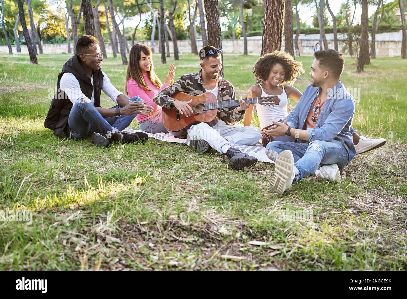 Multi-ethnic friends sitting on the ground while one plays guitar in a ...