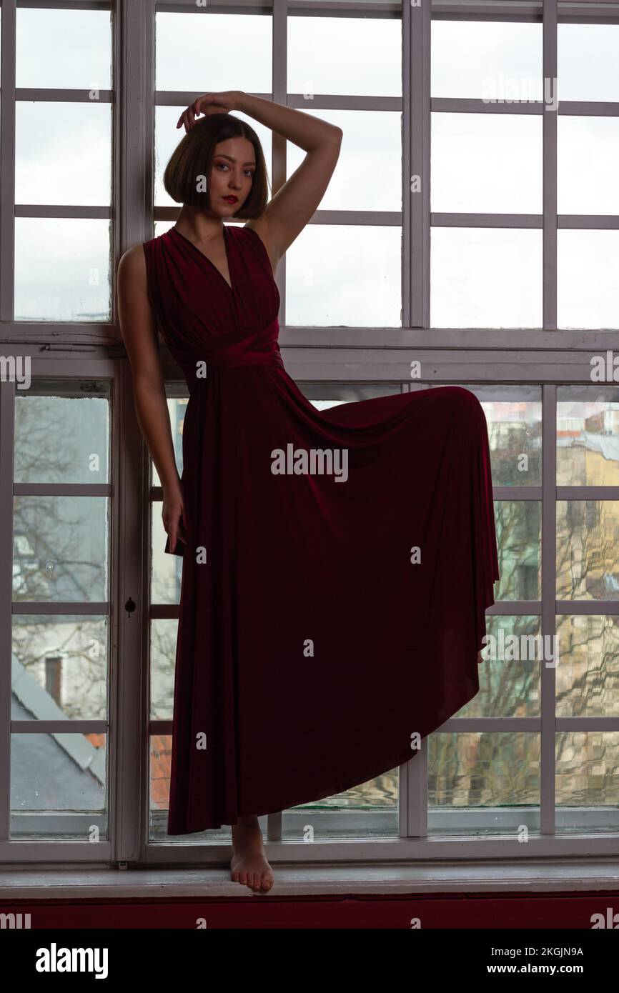 A woman in a red dress with bent leg in front of a small pane window ...