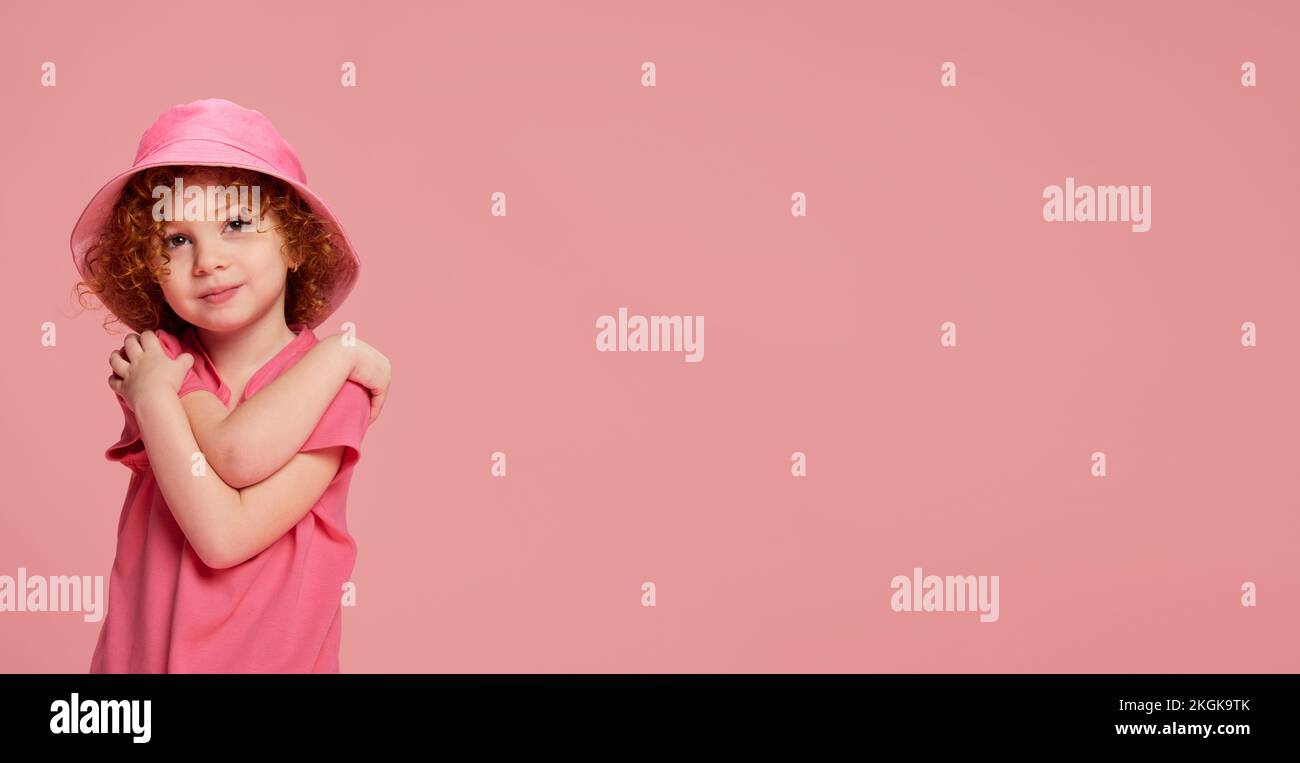 Portrait of cute little girl, child with curly red hair posing in panama isolated over pink ...