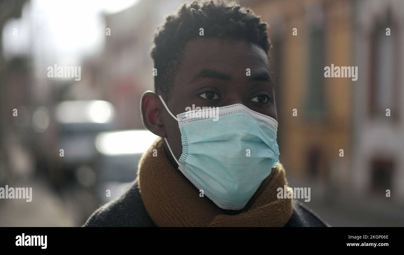 Young black man wearing covid-19 face mask standing outside in sunlight ...