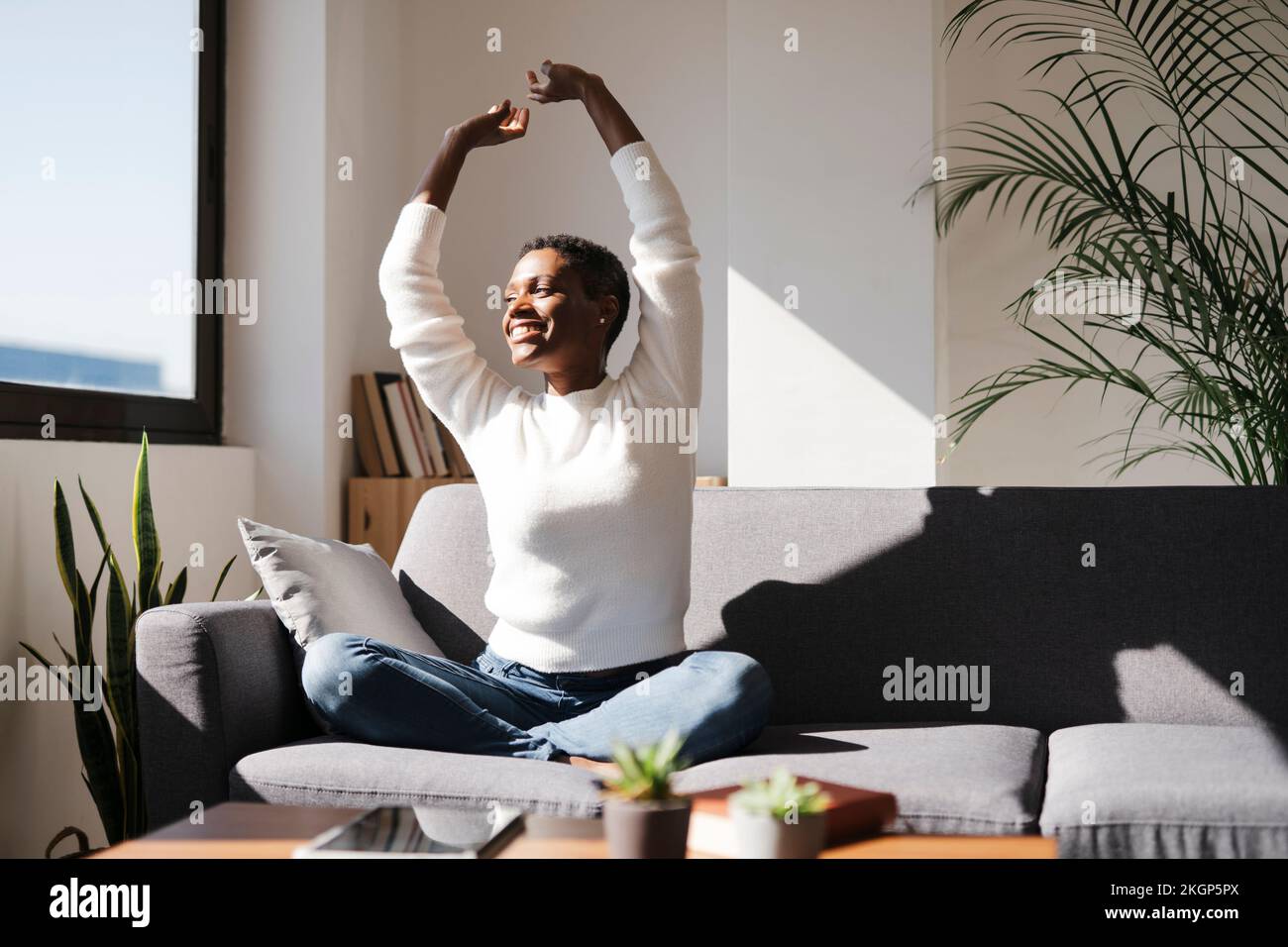 Smiling woman sitting on couch at home stretching her arms Stock Photo ...