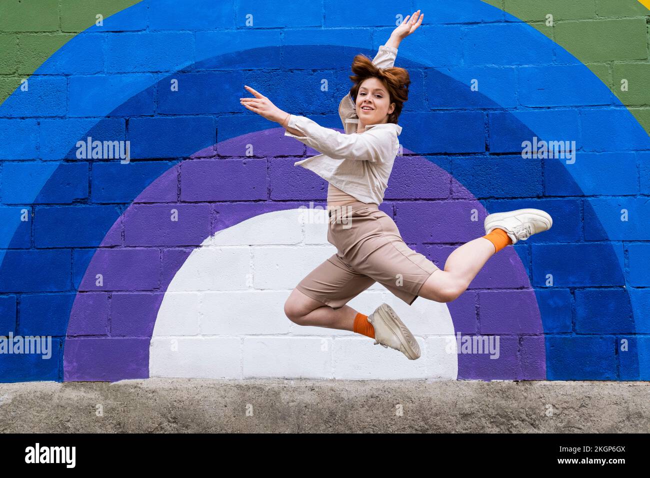 Smiling young woman with hand raised jumping in front of colorful wall ...