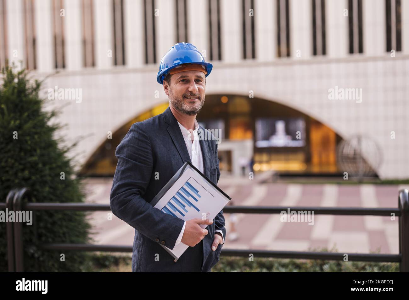 Happy businessman wearing hardhat holding chart in front of railing ...