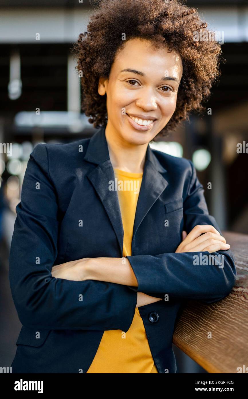 Happy businesswoman wearing blazer leaning on railing with arms crossed ...