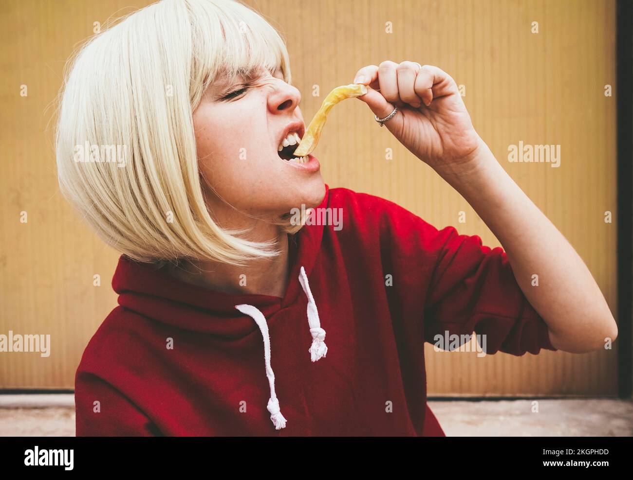 Blond woman eating French fries in front of brown wall Stock Photo - Alamy
