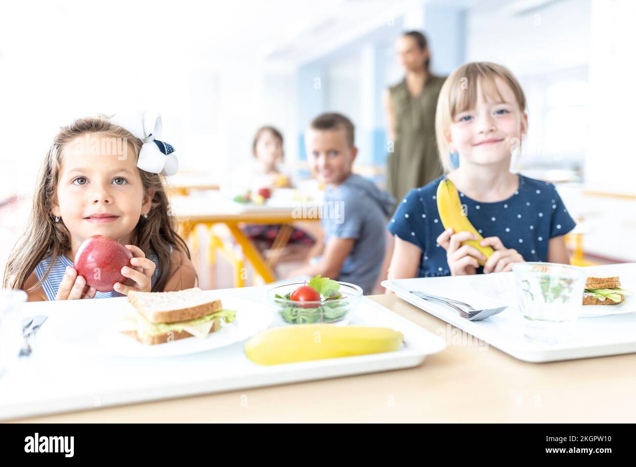 Girls holding apple and banana sitting at table in cafeteria Stock ...