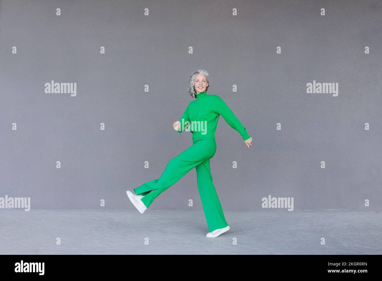 Smiling woman standing on one leg in front of gray wall Stock Photo - Alamy