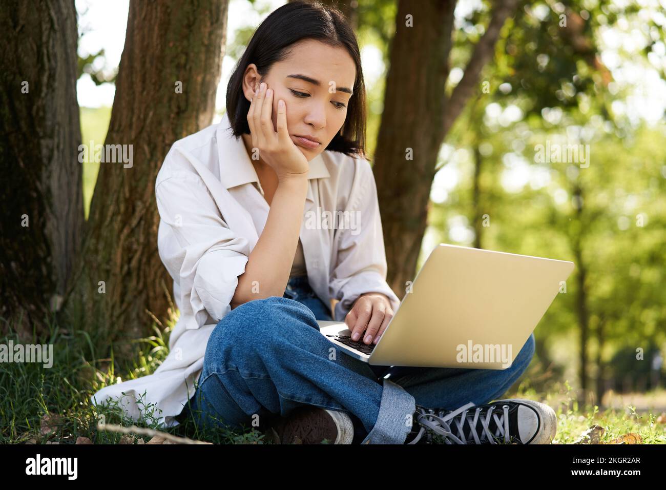 Portrait Of Asian Woman Sitting On Grass Near Tree Using Laptop