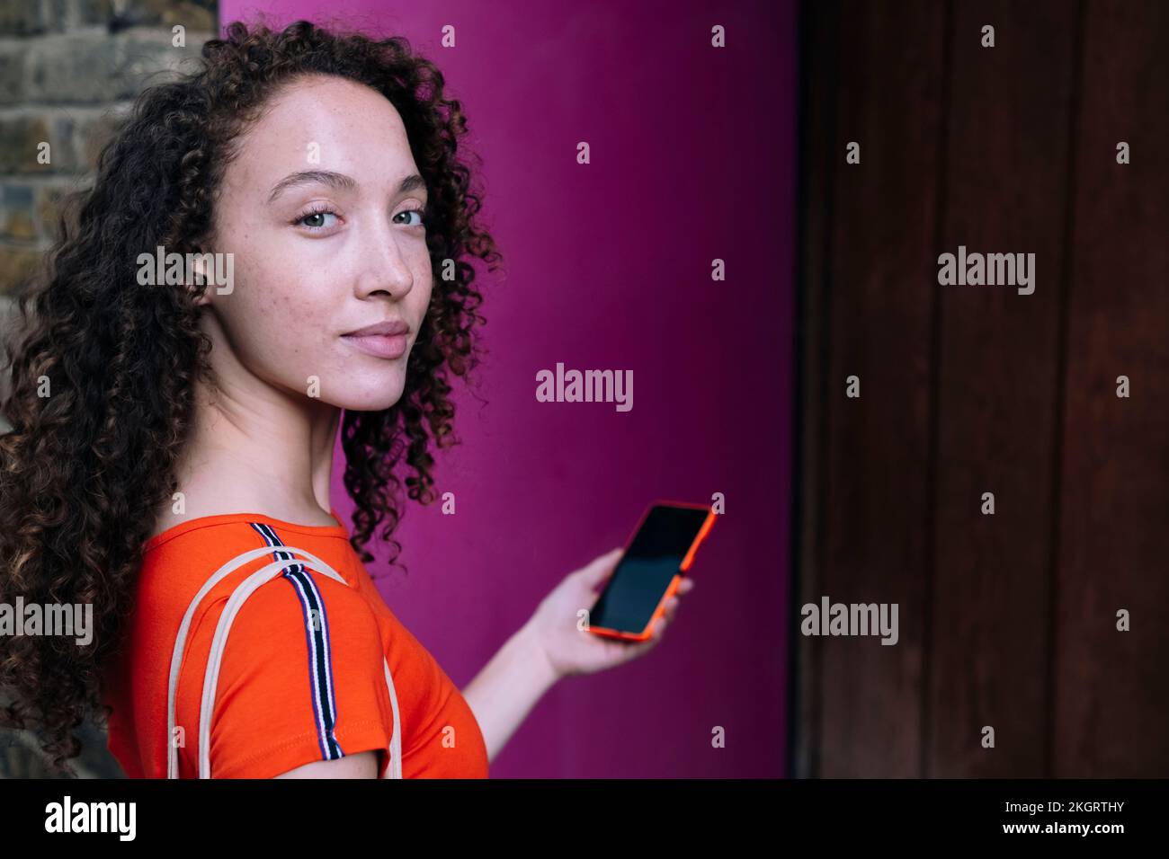 Young woman with mobile phone standing by wall Stock Photo - Alamy