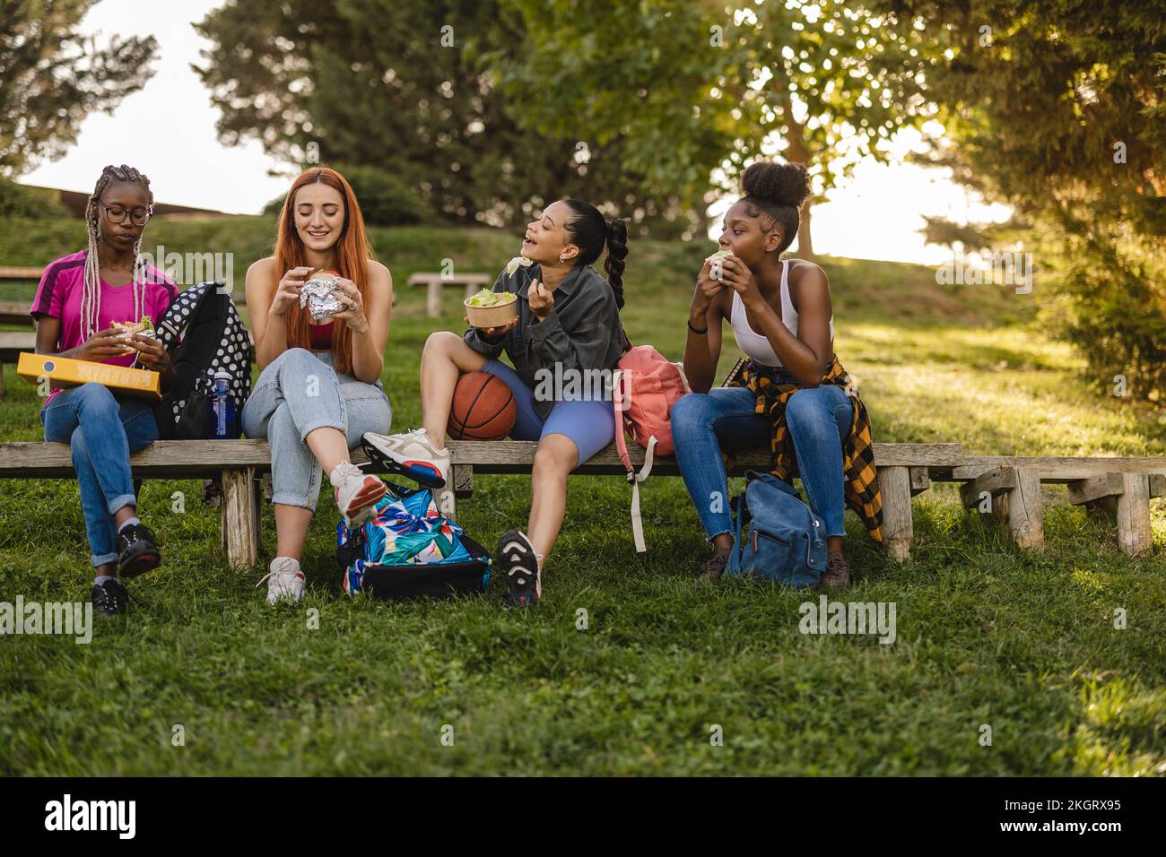 Happy friends sitting on bench enjoying food in park Stock Photo - Alamy