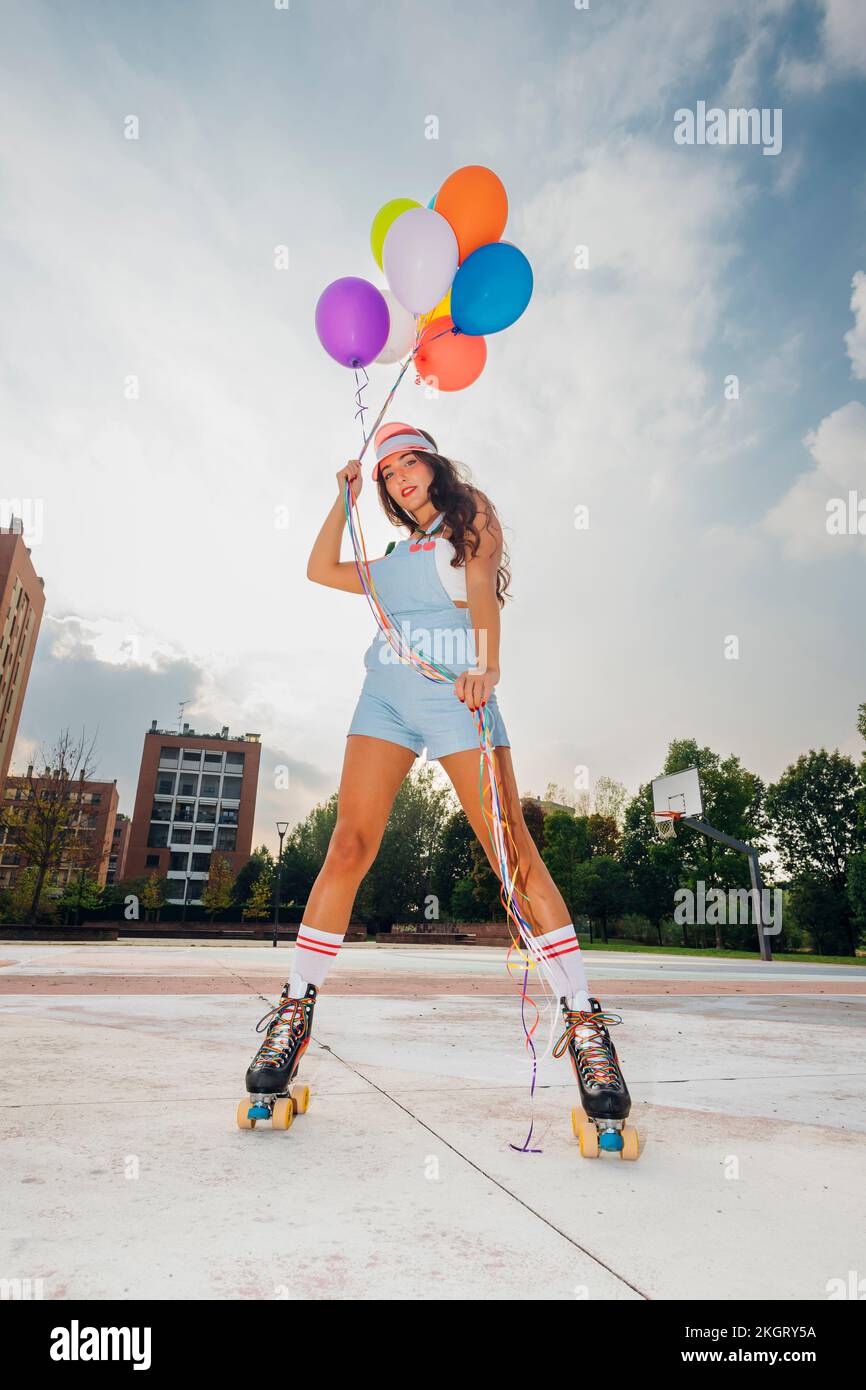 Young woman wearing roller skates holding colorful balloons under sky ...