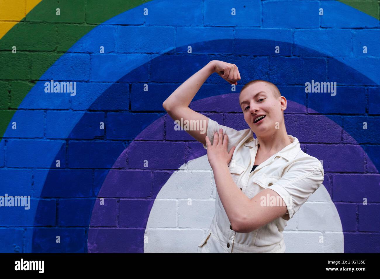 Non-binary person flexing muscles in front of multi colored wall Stock ...