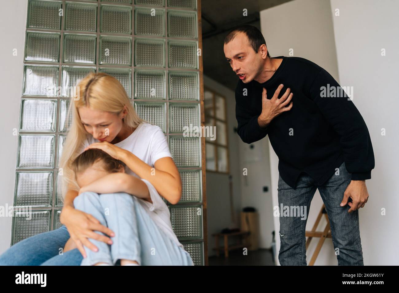 Scared mother and daughter hugging each other sitting on sofa while aggressive standing father ...