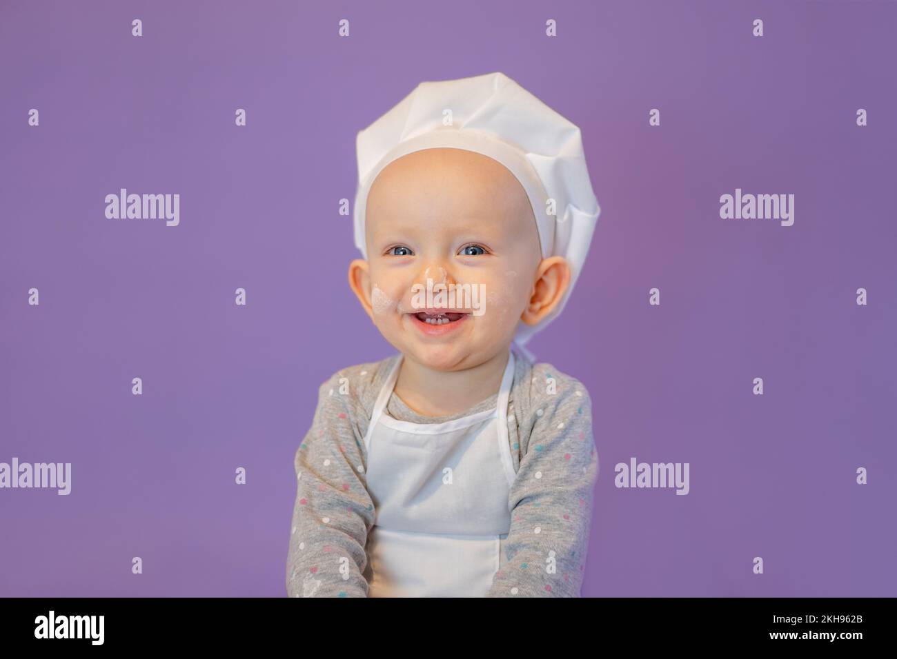 little cute baby in an apron and a chef's hat smiles on a lilac ...