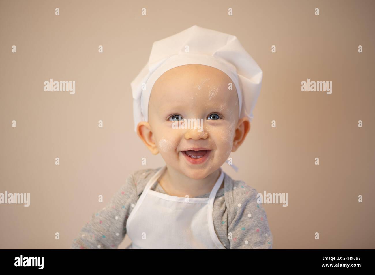 little cute baby in an apron and a chef's hat smiles on a beige ...