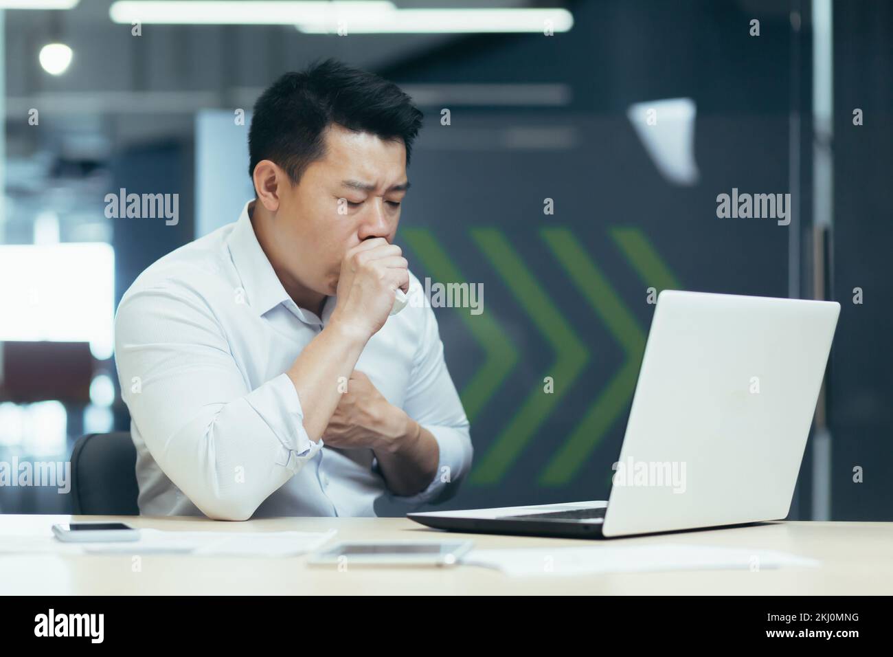 A young Asian freelancer, an office worker fell ill at work. He sits at the table and coughs ...