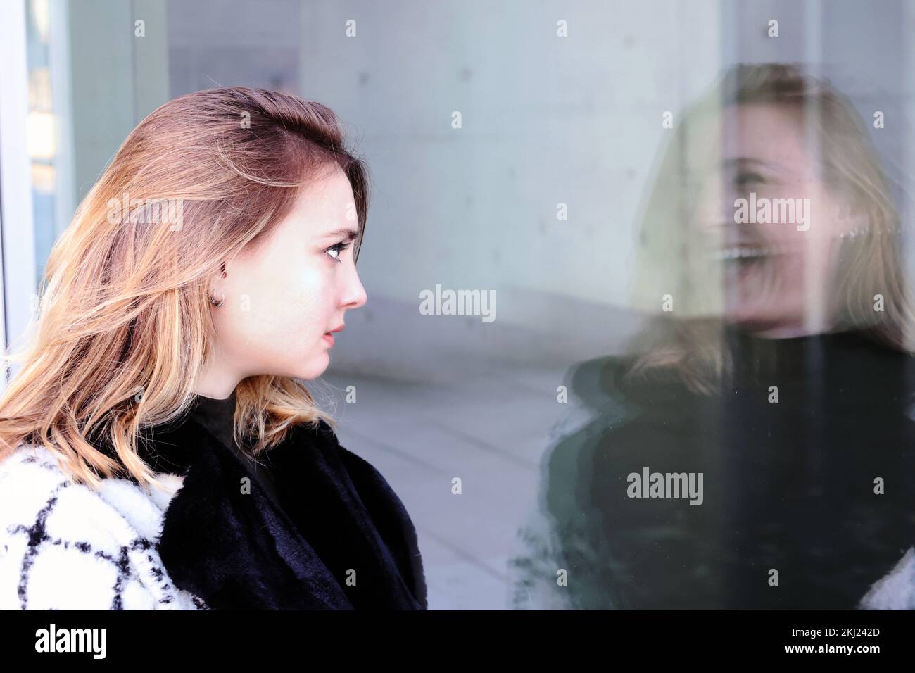 young woman standing outside and looking at her laughing reflection at ...