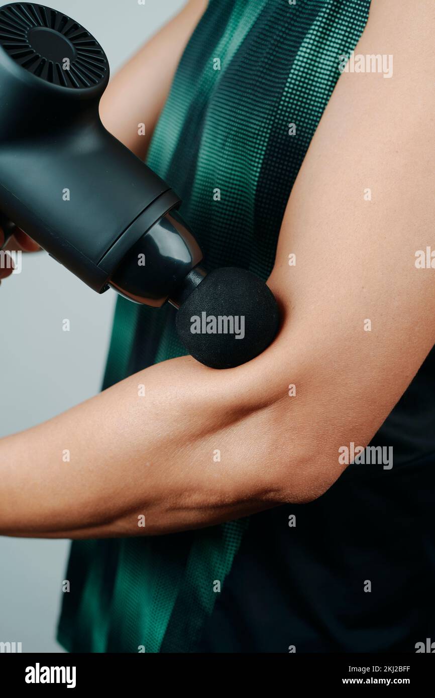 a young caucasian man, in sportwear, uses a massage gun to massage the ...