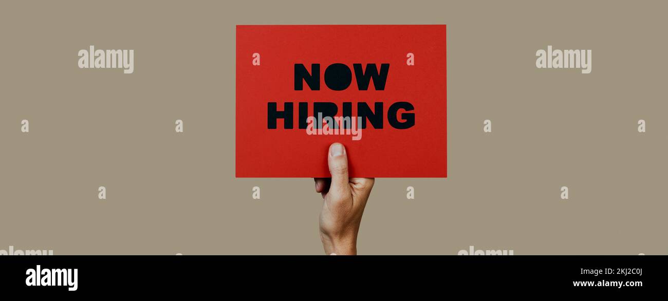 a man holds a red paper sign with the text now hiring written in black ...