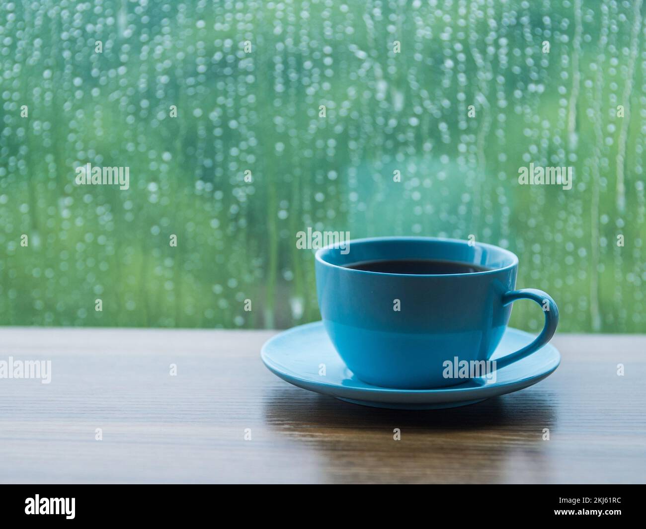 A blue cup of coffee on a wooden table in front of a window with water ...