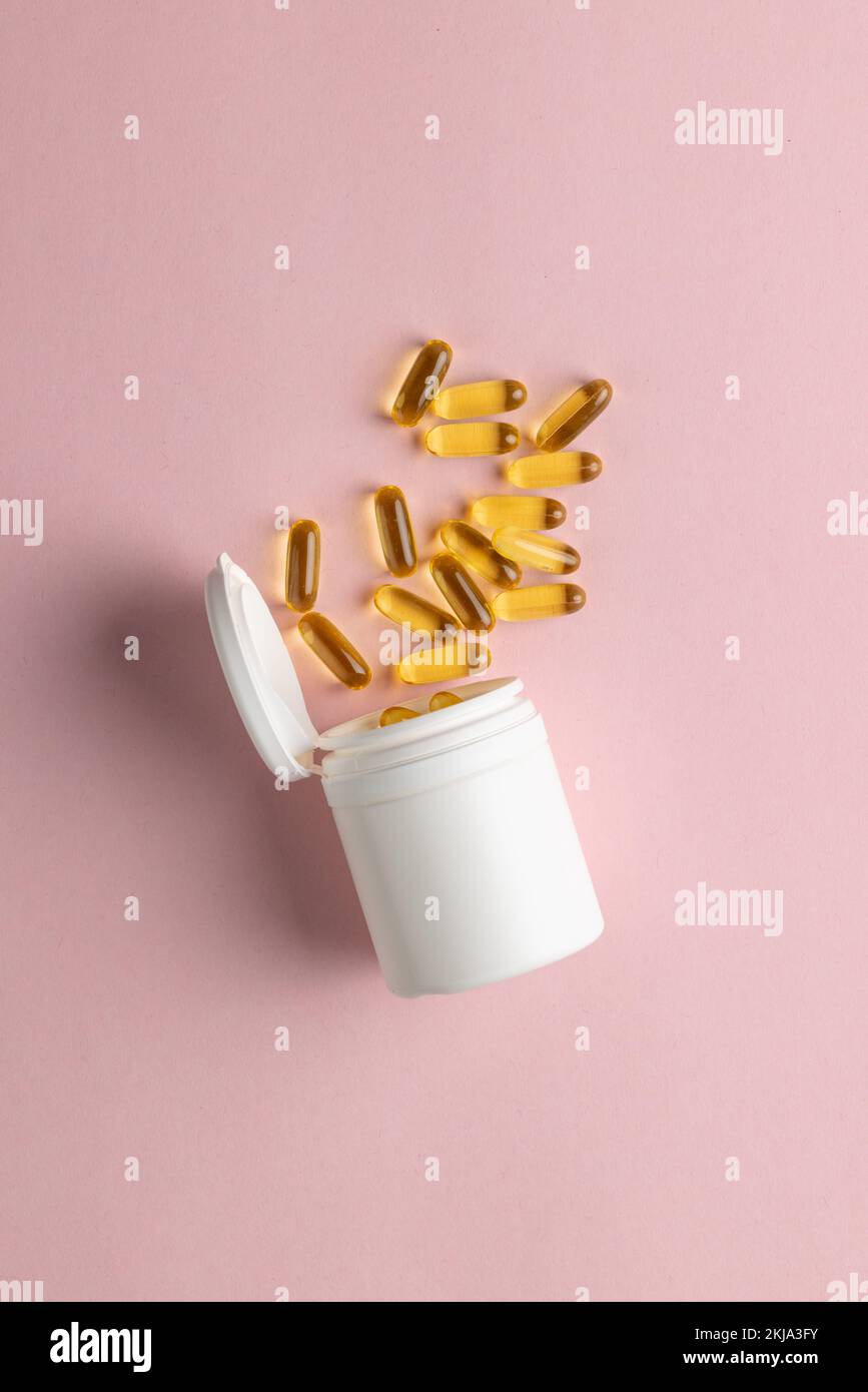 Vertical composition of white pill box spilling oil capsules on pink ...
