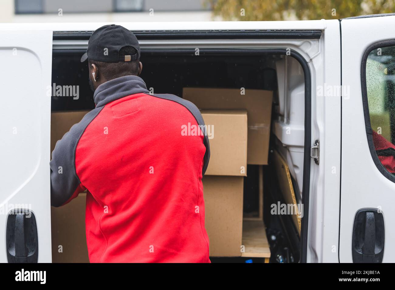 Black young adult delivery man in work uniform unloading cardboard box packages from white van ...