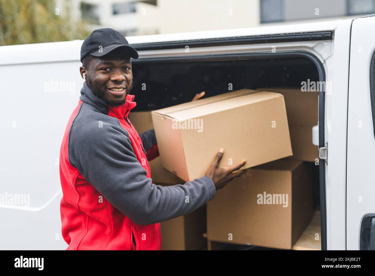 Black young adult delivery guy in work uniform and black cap unloading packages from white van ...