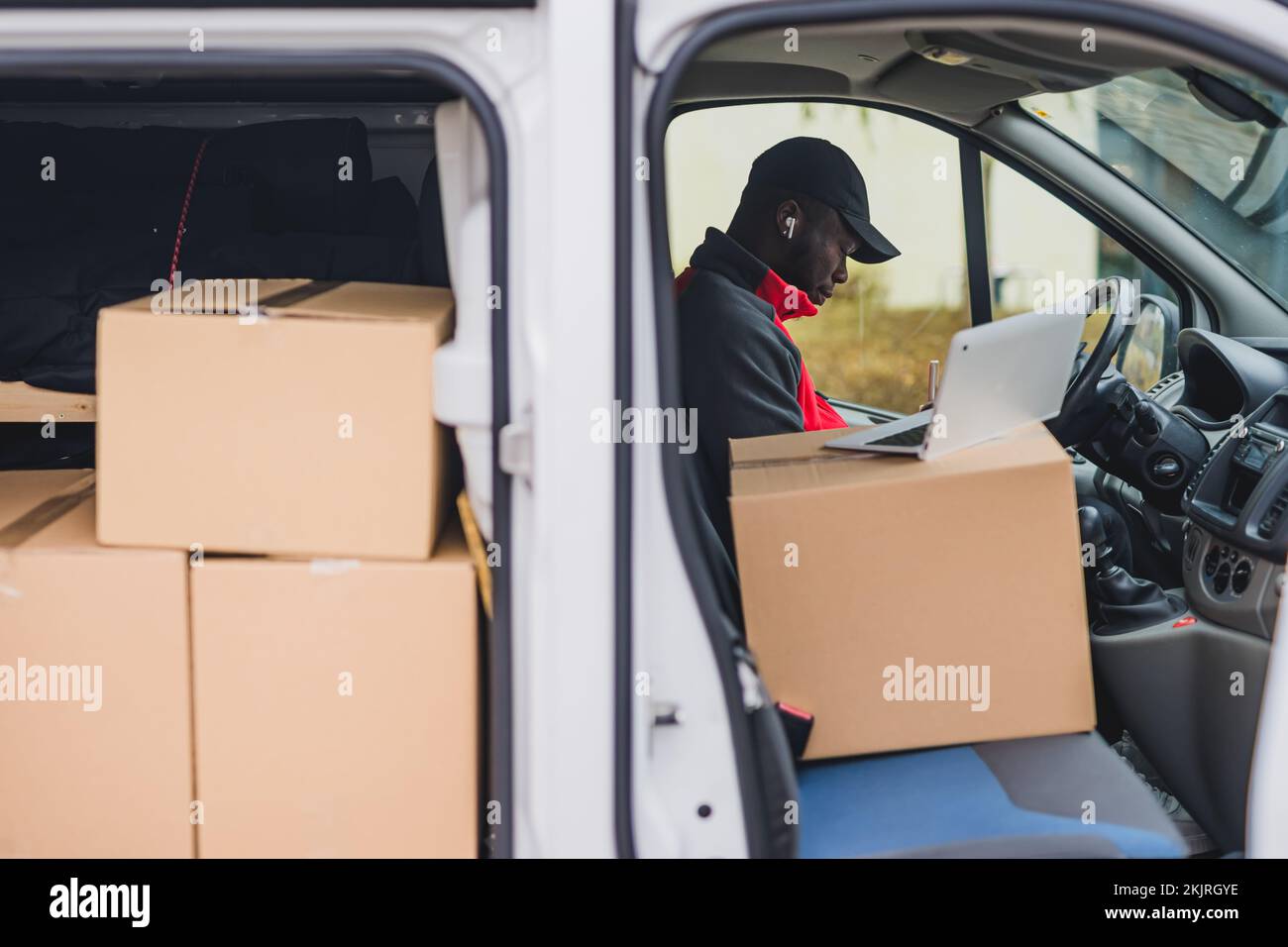 Black young adult delivery man sitting in white van with cardboard box packages filling out ...