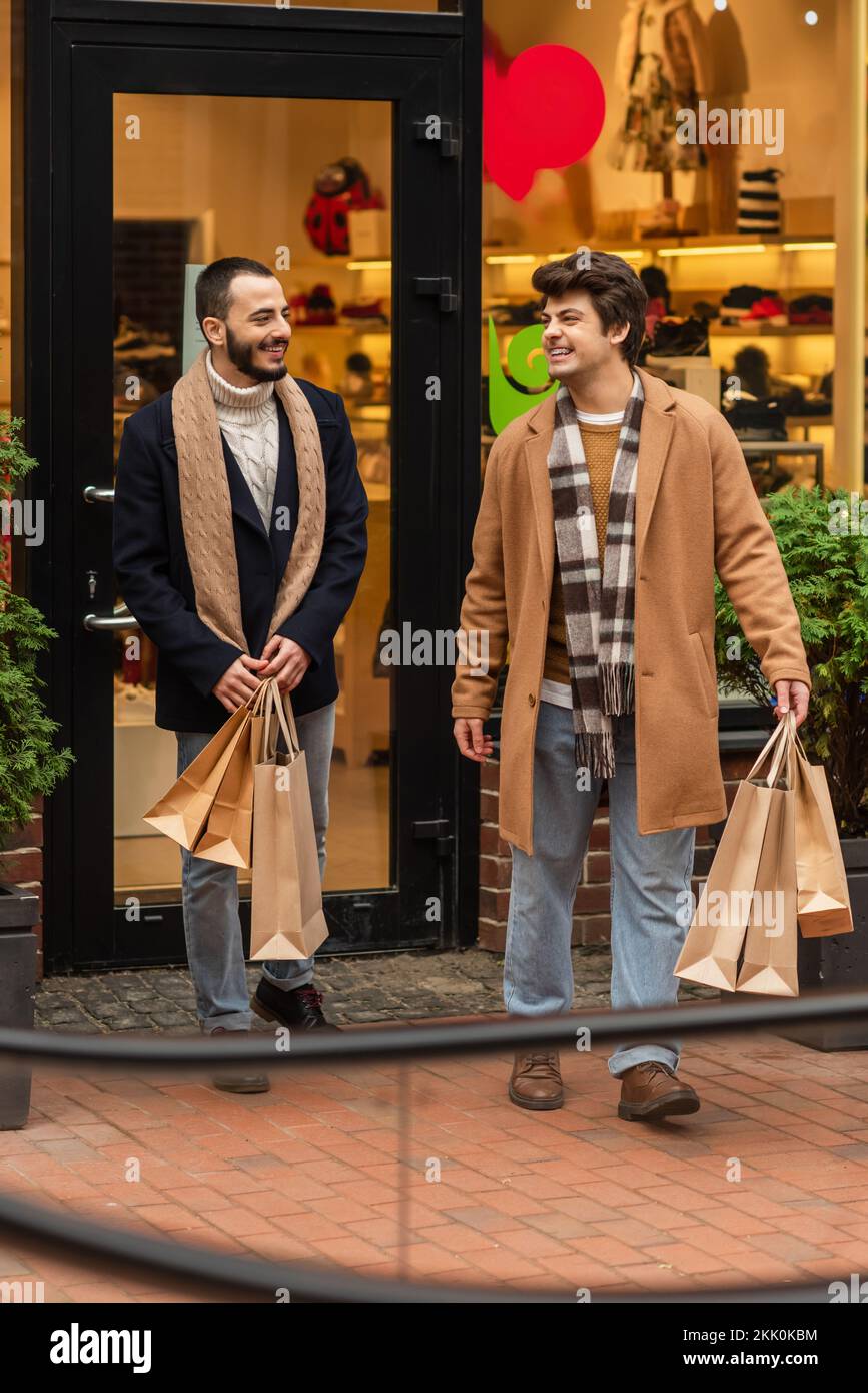 full length of cheerful gay men in stylish clothes holding shopping ...