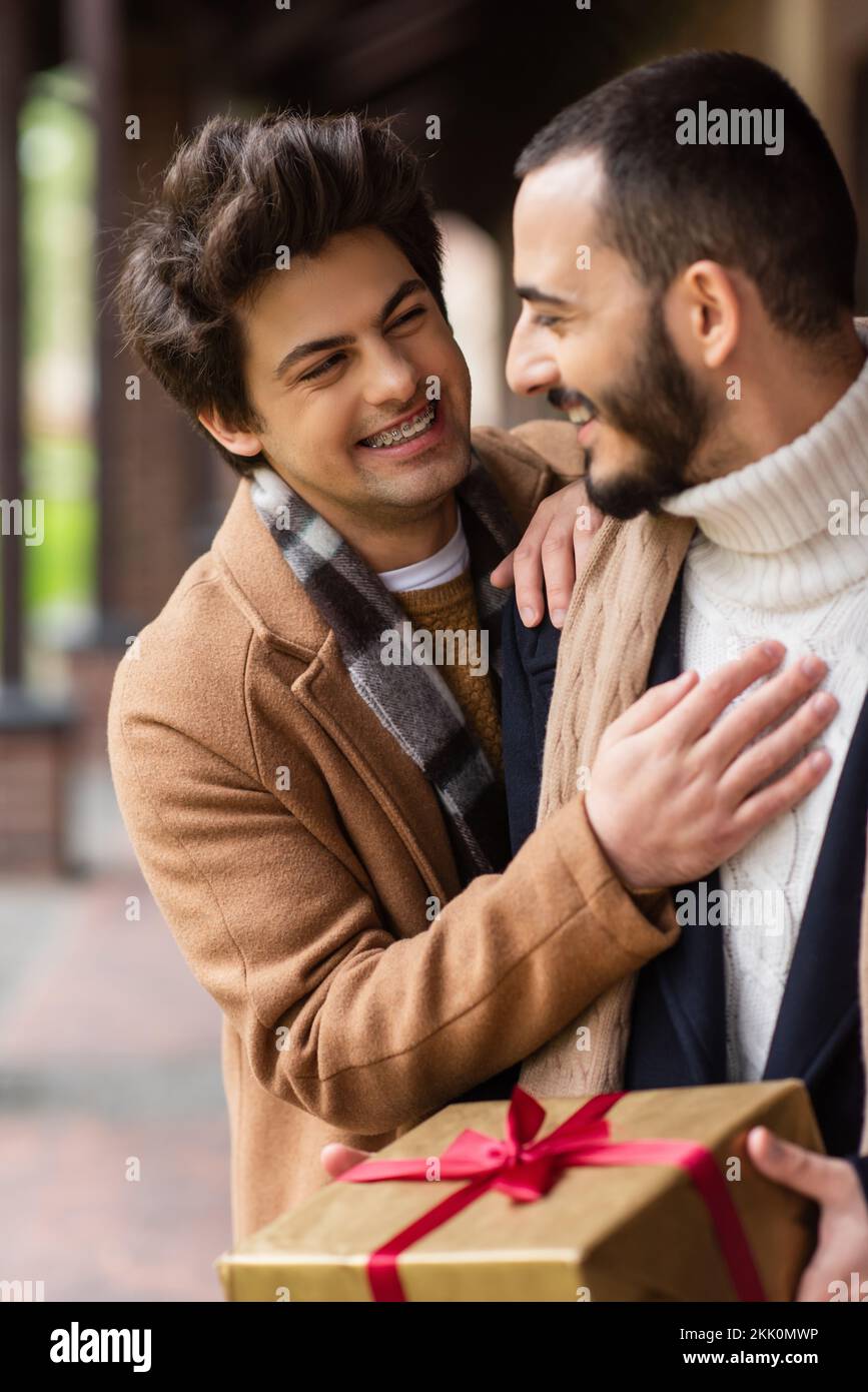young and cheerful gay man touching chest of bearded boyfriend holding ...