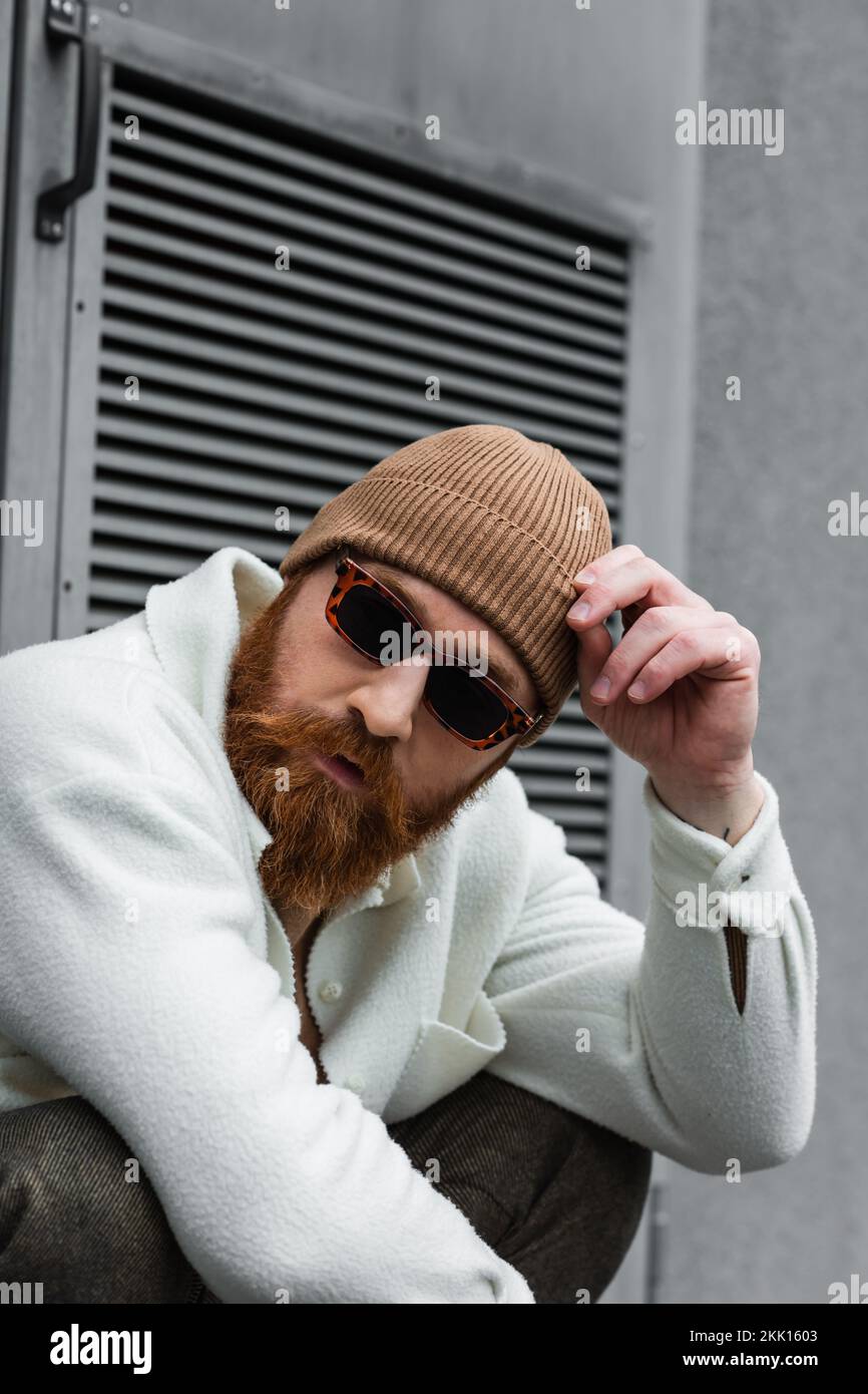 bearded man in stylish sunglasses and trendy beanie hat looking away ...