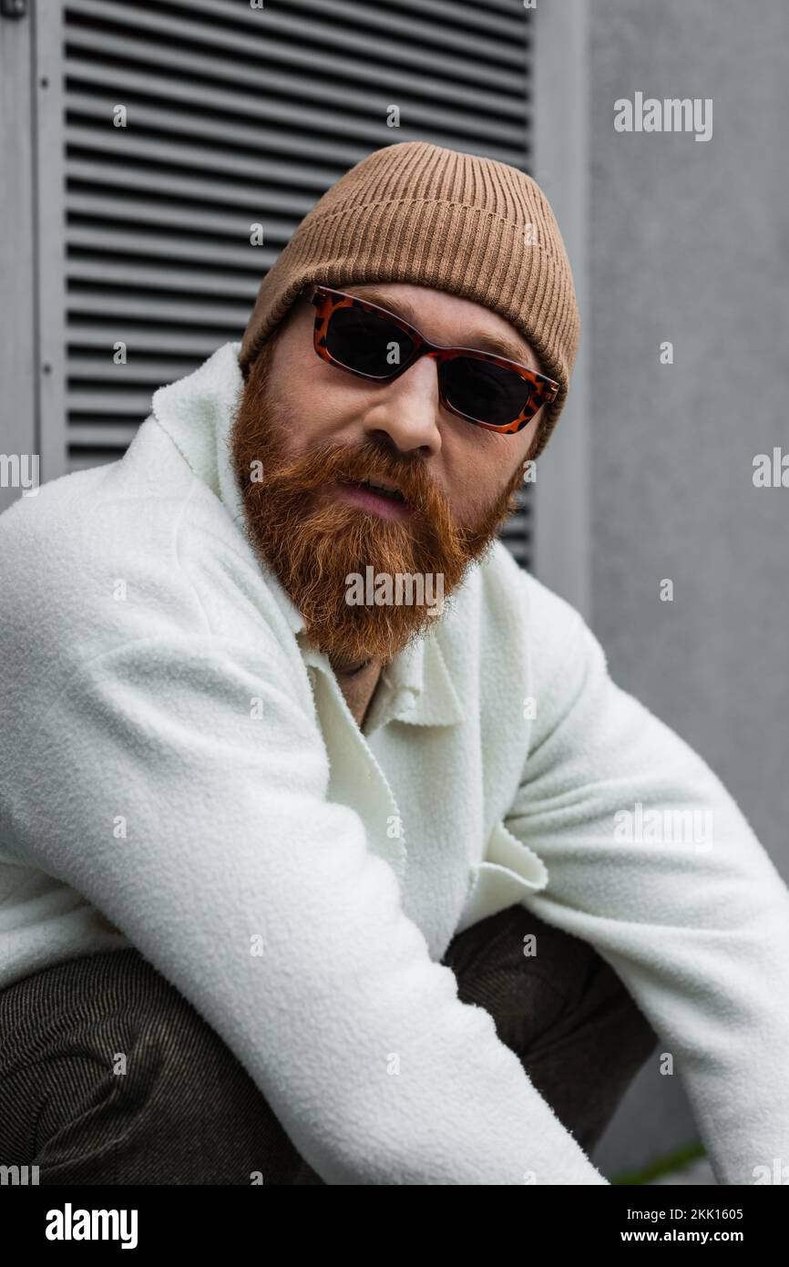 bearded man in stylish sunglasses and beanie hat looking away outdoors ...