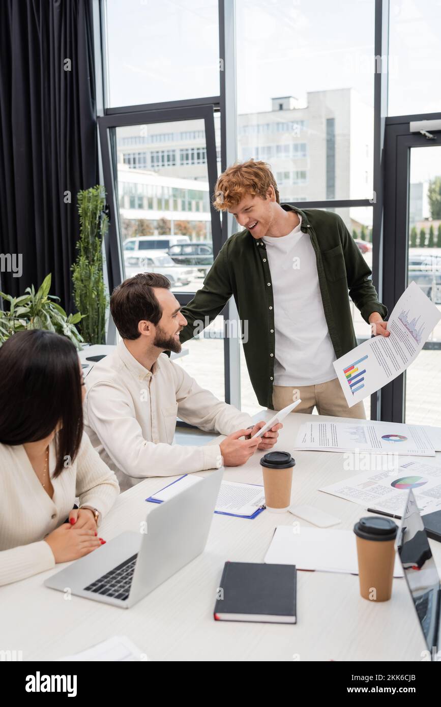 smiling man with infographics talking to interracial colleagues in ...