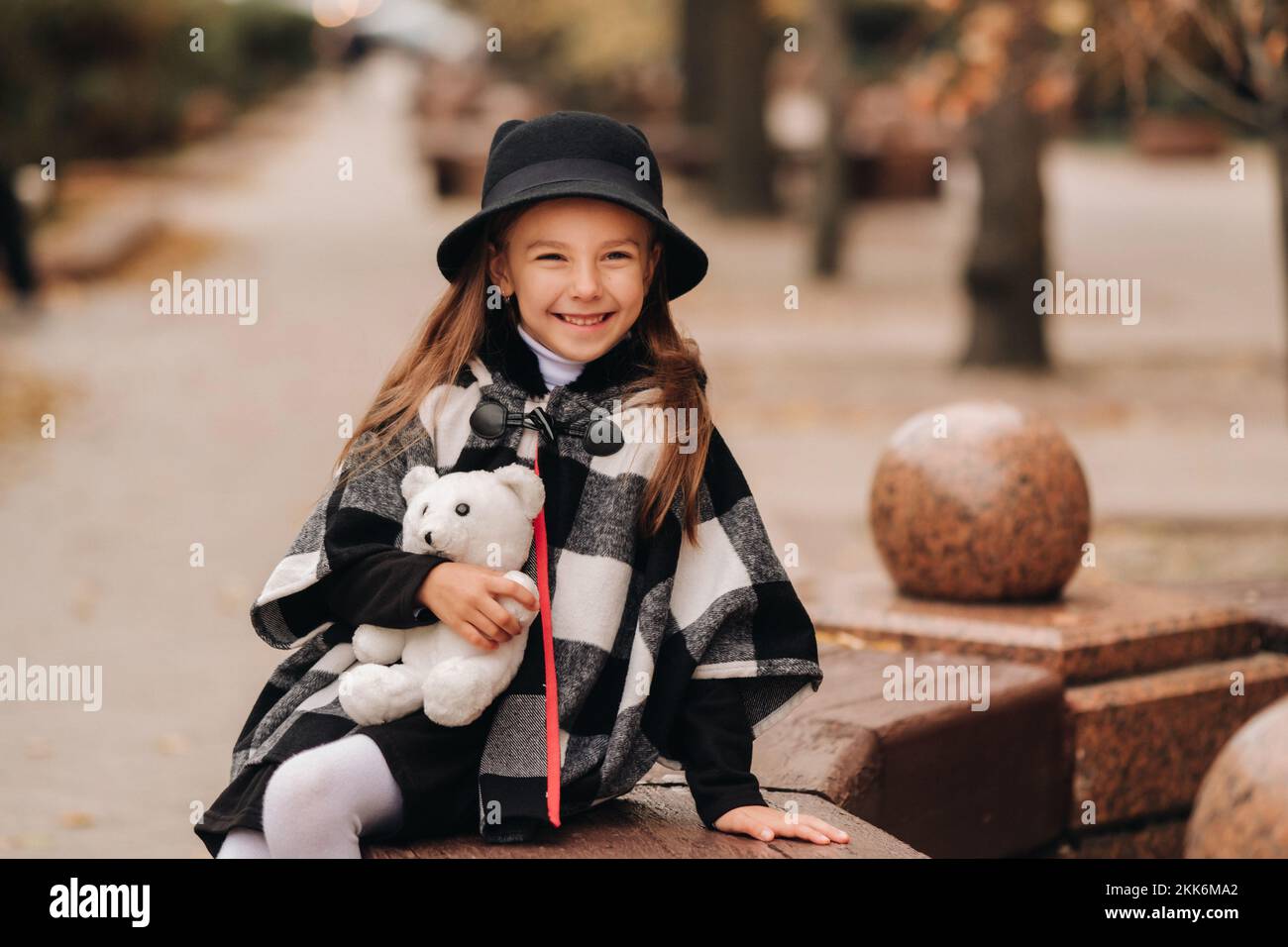 A little stylish girl in autumn clothes is sitting on a bench in the ...