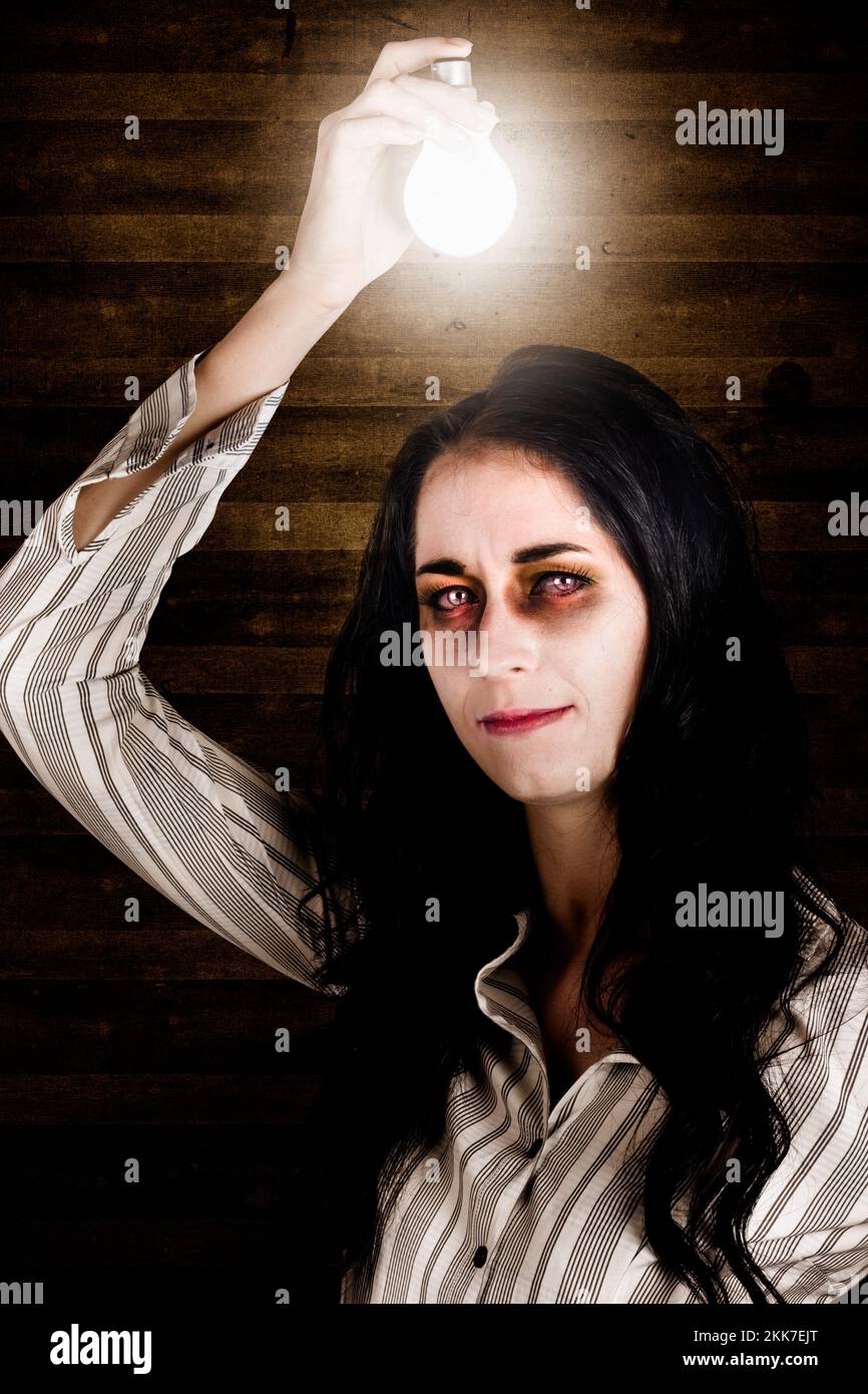 Creepy attic girl wearing devilish makeup holding a lit bulb above her ...