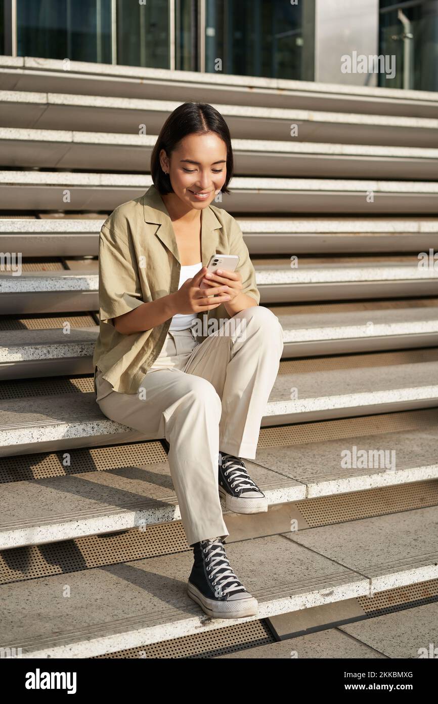 Beautiful asian girl sitting on stairs outside building, using mobile ...