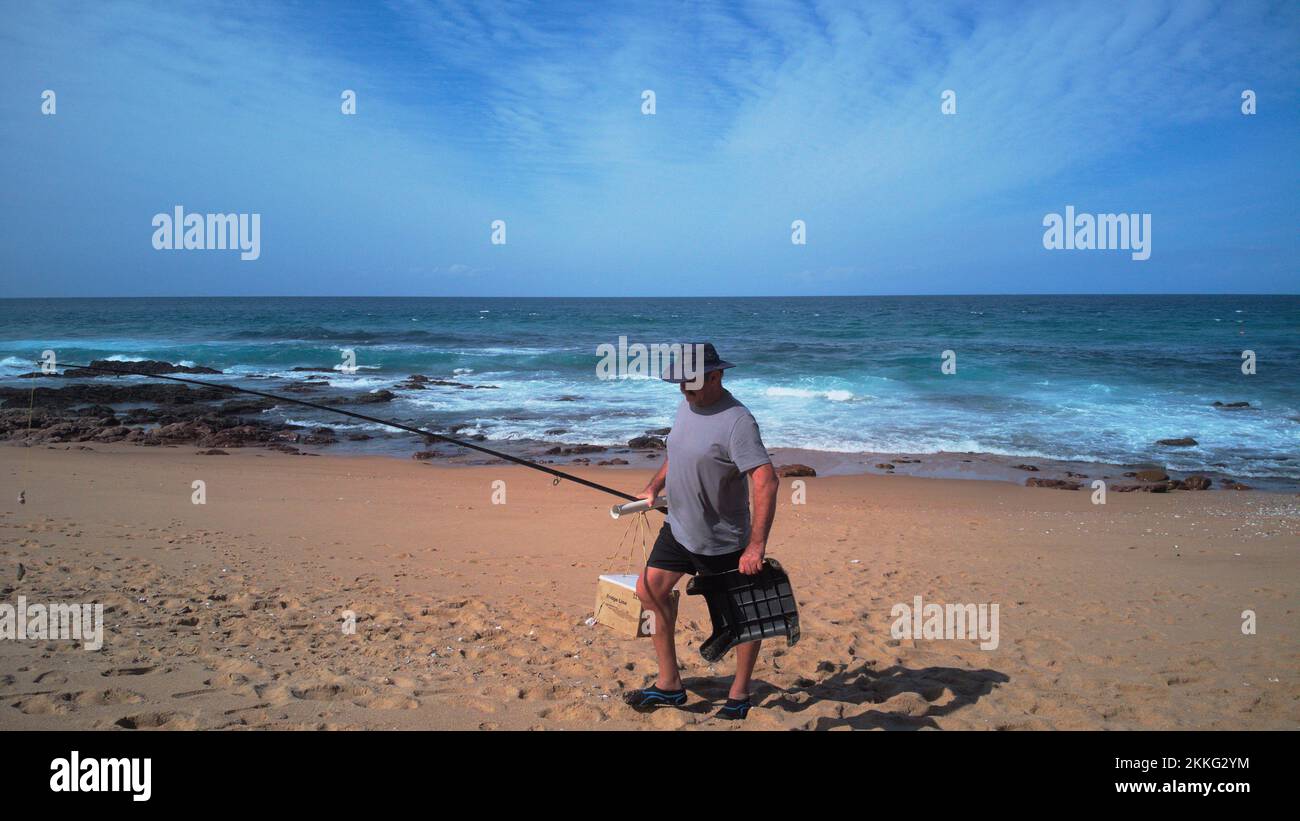 A man with a fishing rod walking on the sands of a beach by a bright ...