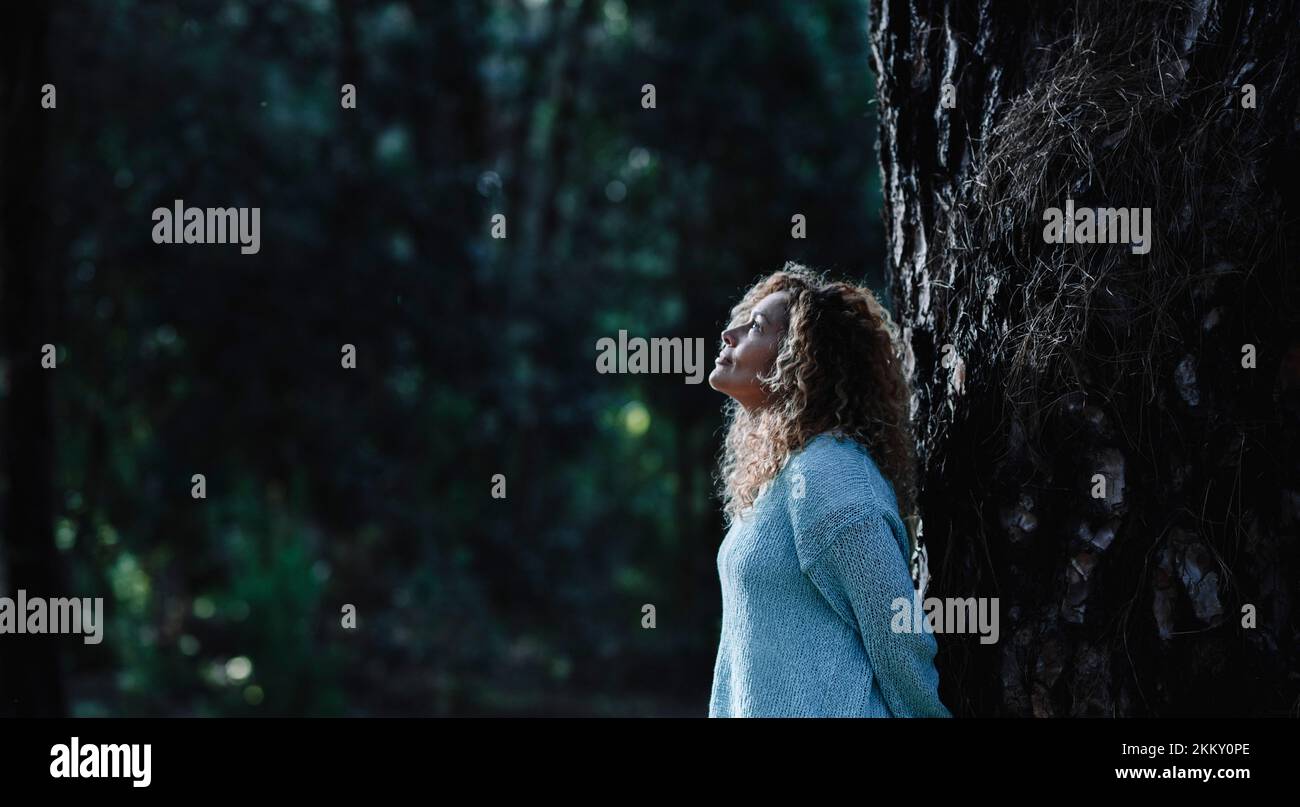 Thoughtful woman standing against a trunk tree in the forest looking up ...