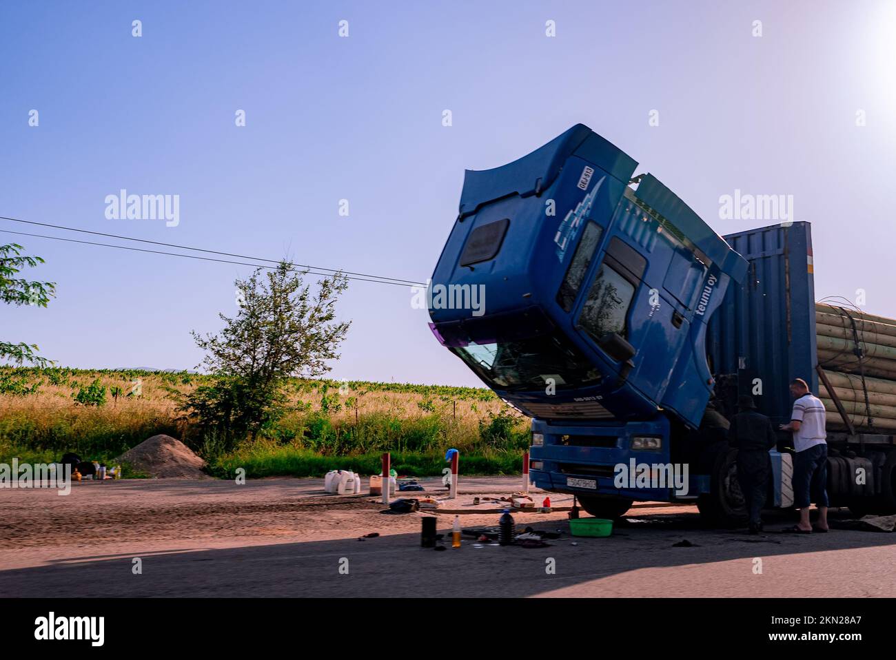 A blue broken-down truck on the side of a road with two men trying to ...