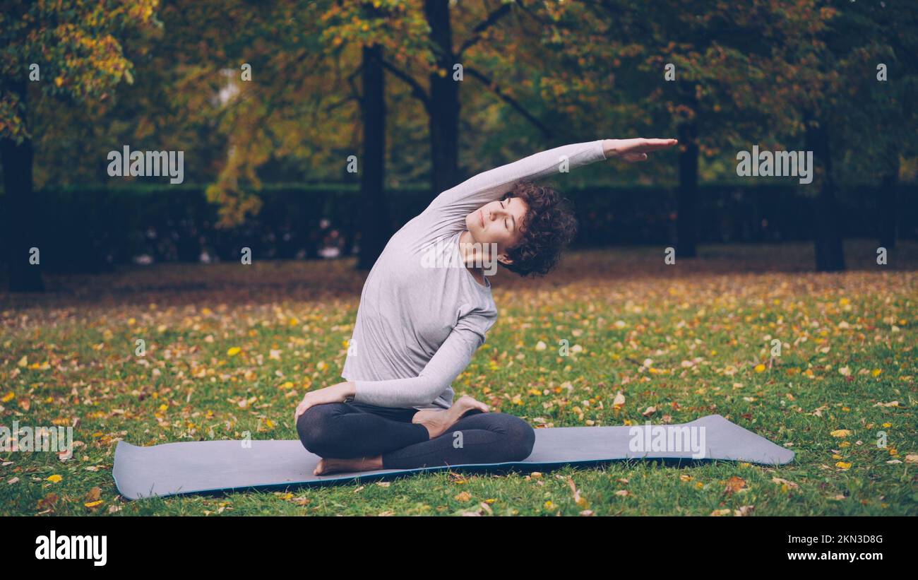 Slim young lady is doing sequence of yoga asanas sitting on mat in park ...