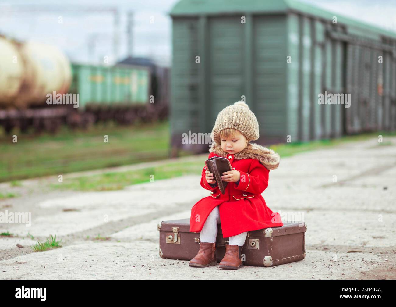 charming baby in a red coat with a suitcase is waiting for a train ...