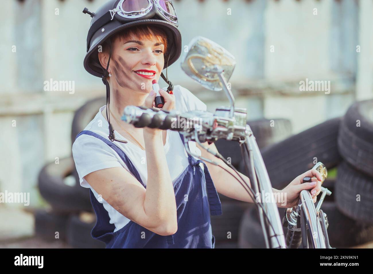 Girl auto mechanic in helmet looks in motorcycle mirror and paint lips ...