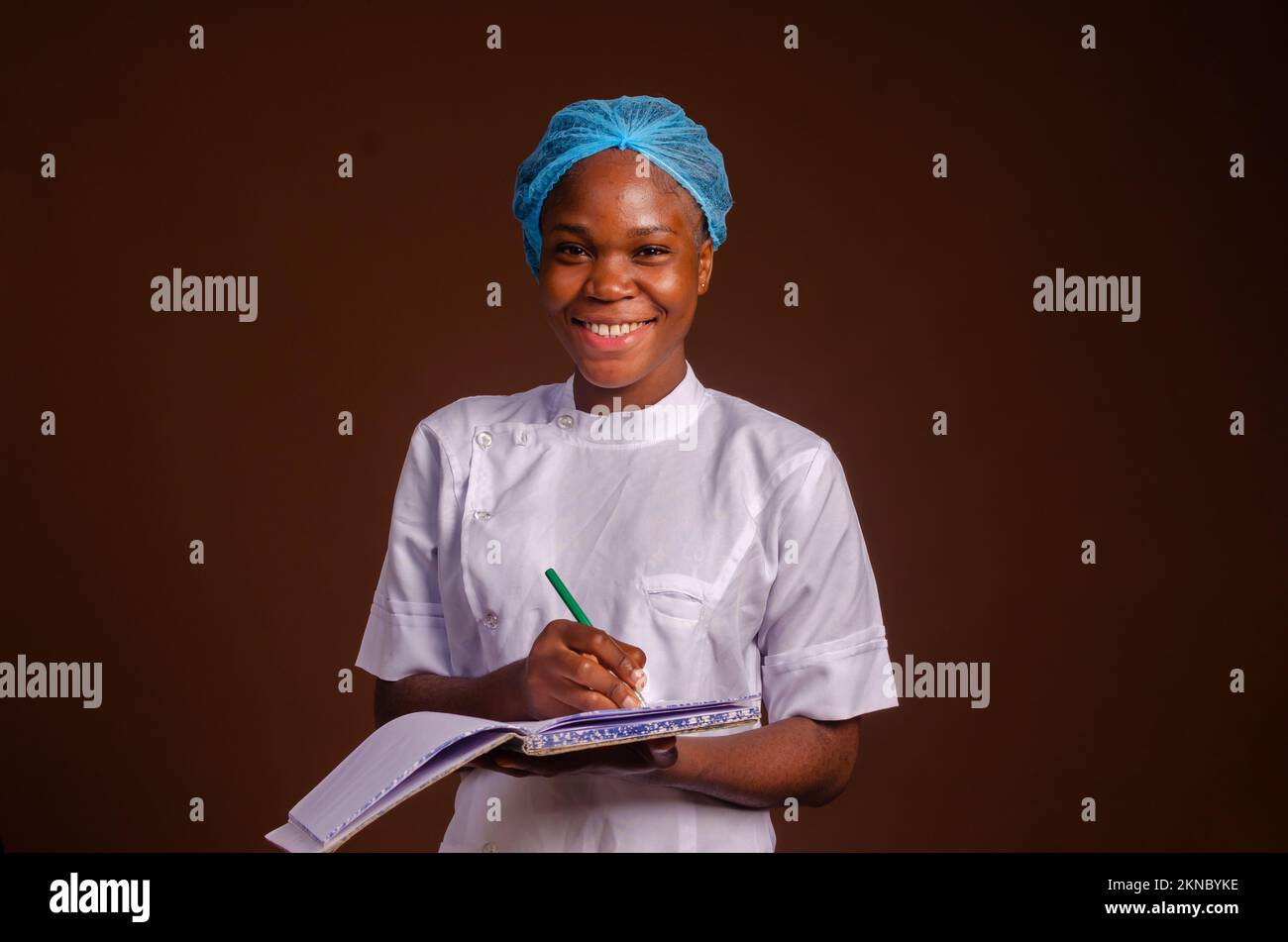 A smiling Nigerian female medic writing on a book isolated on a brown ...