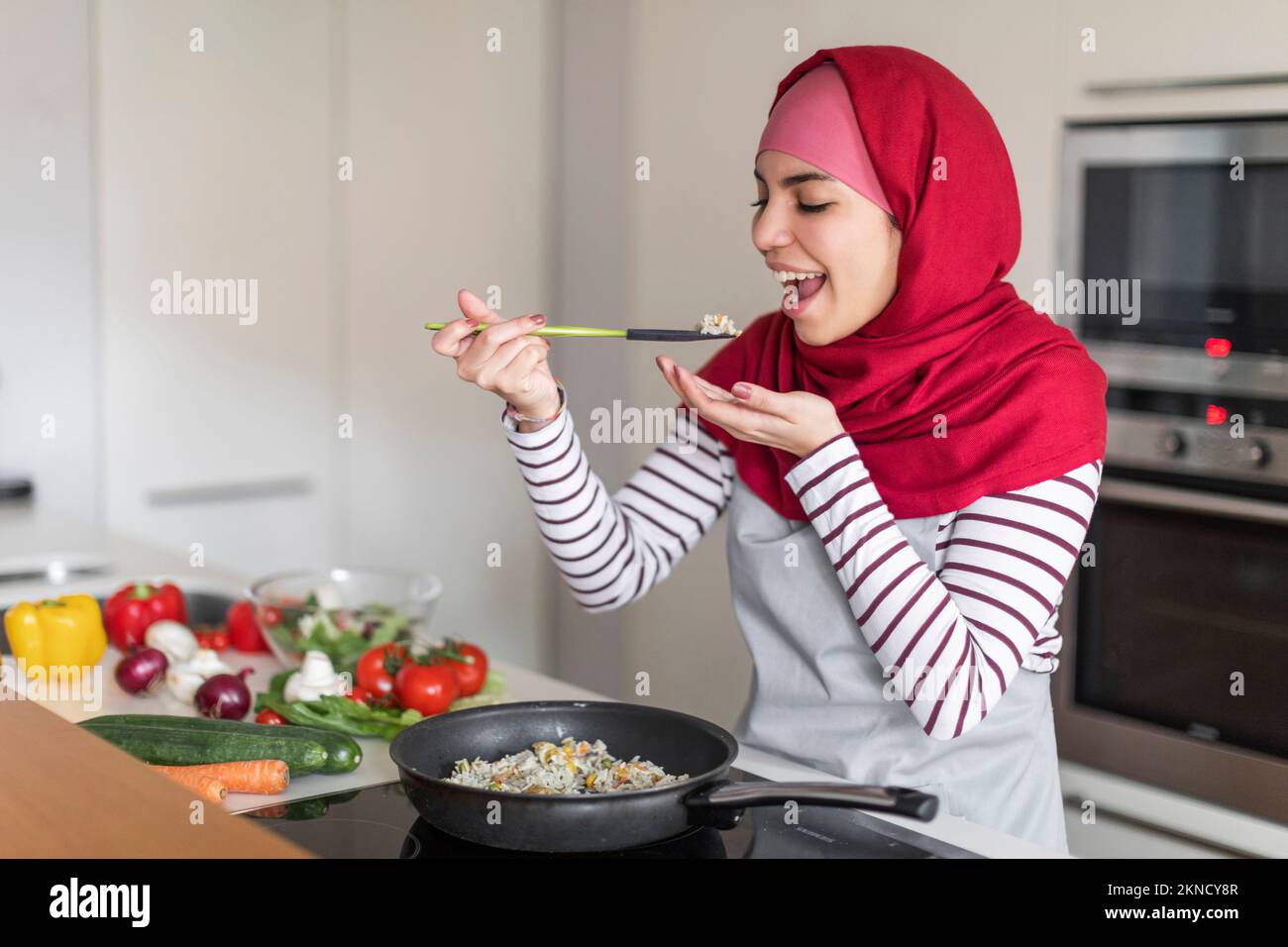 Cheerful arab woman housewife cooking on electric stove, tasting food ...