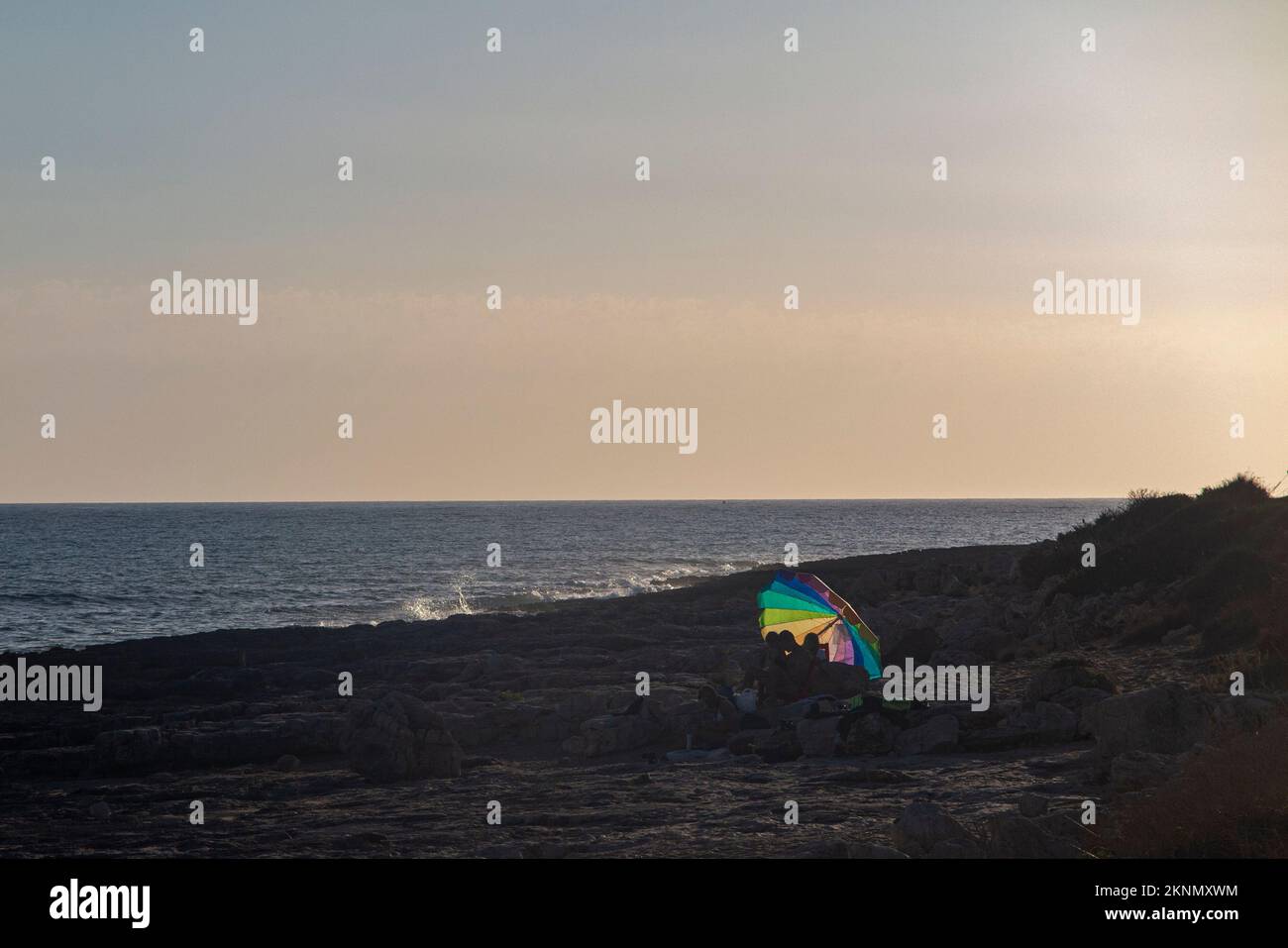 Protection from the sun under a rainbow coloured beach umbrella backlit from the afternoon sun ...
