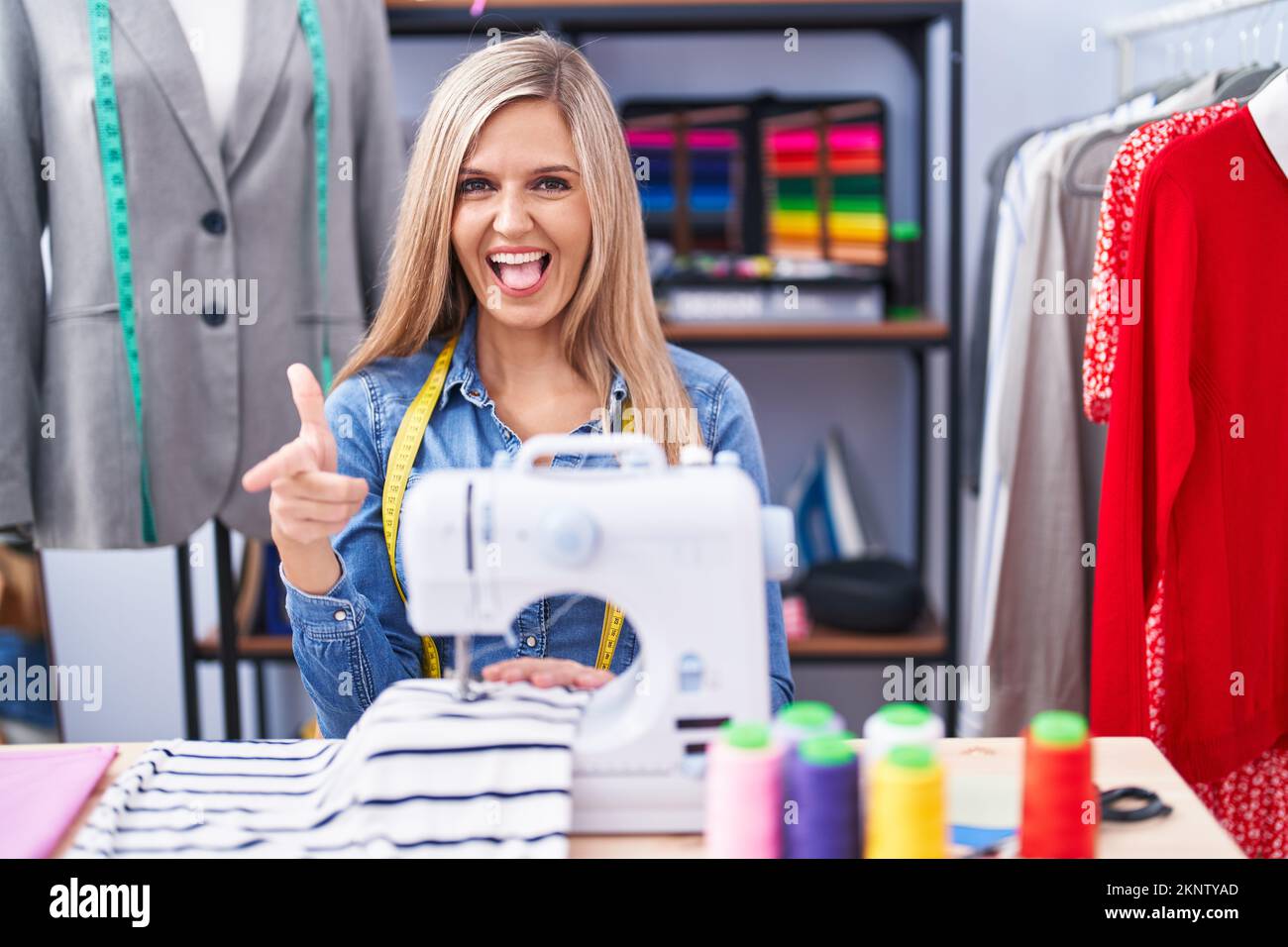 Blonde woman dressmaker designer using sew machine pointing fingers to ...