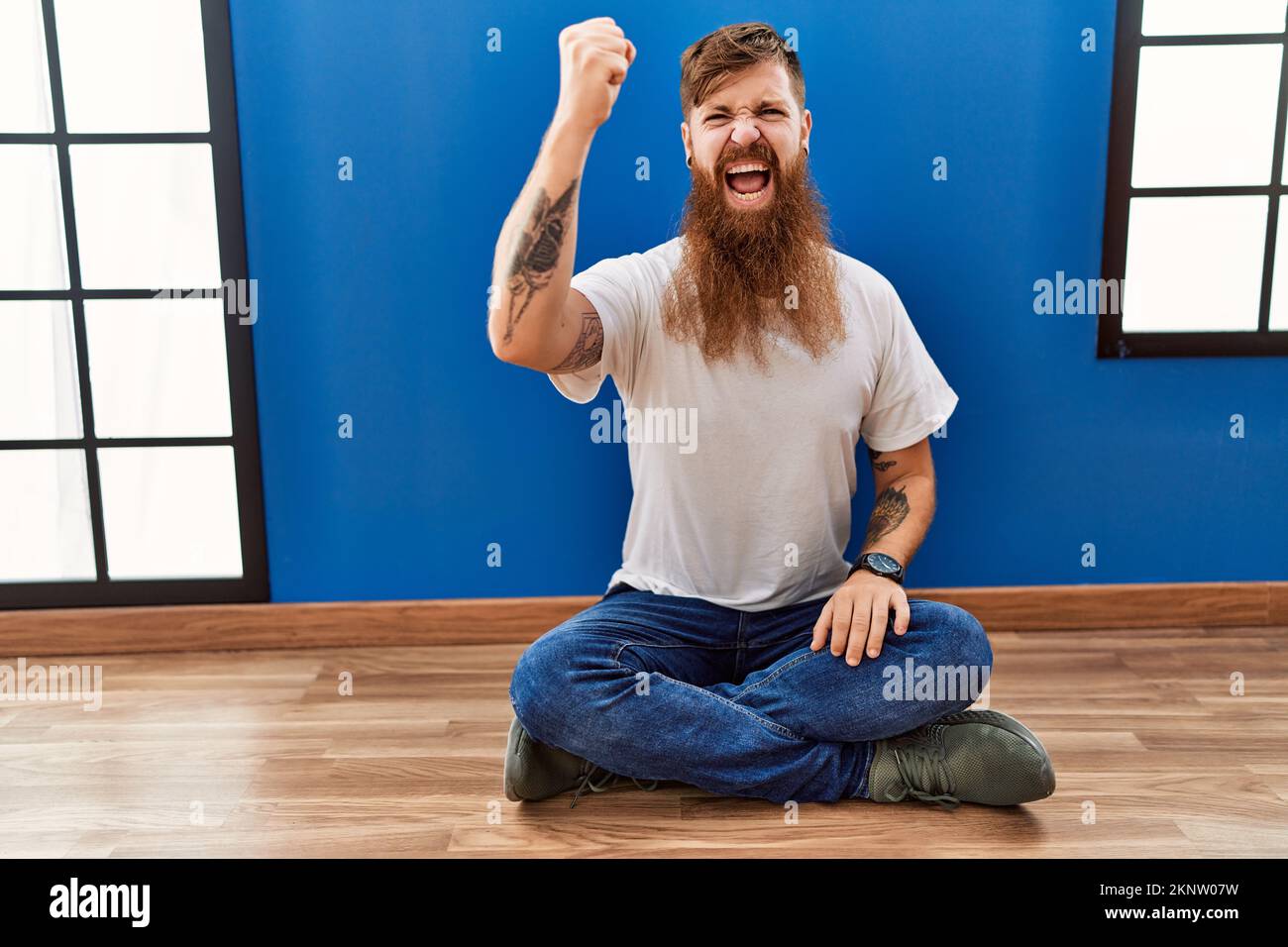 Redhead man with long beard sitting on the floor at empty room angry ...