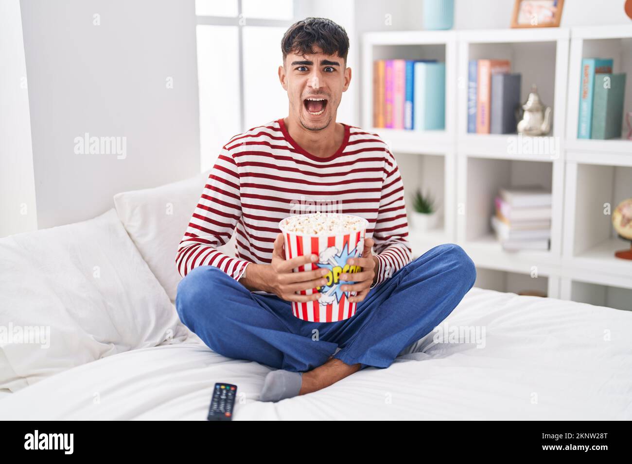 Young hispanic man eating popcorn sitting on the bed watching a movie ...