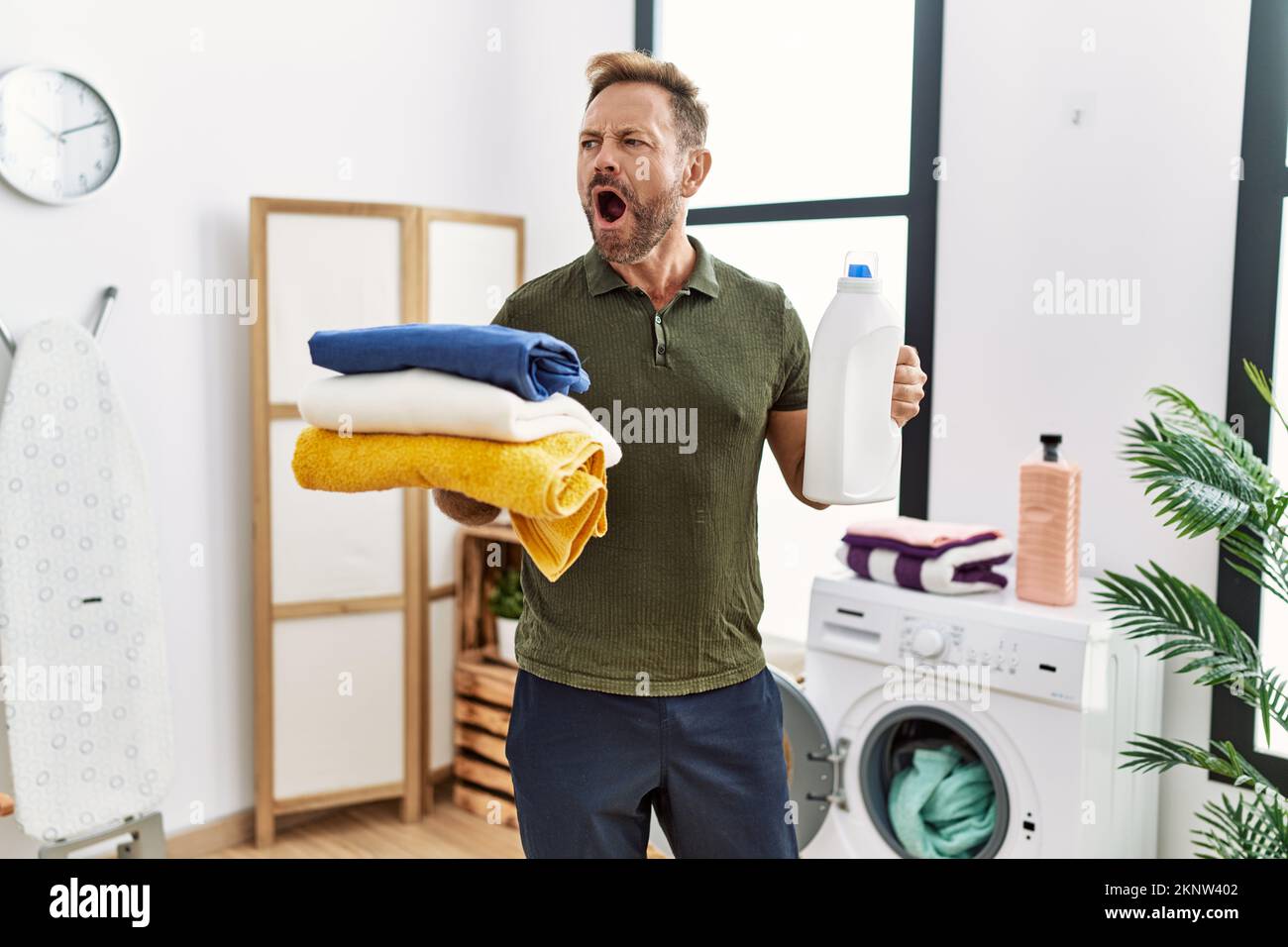 Middle age man holding clean laundry and detergent bottle angry and mad ...