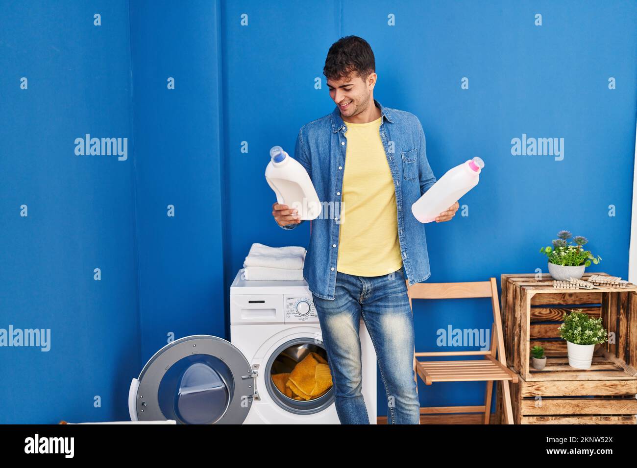 Young hispanic man washing clothes holding detergent bottles at laundry ...